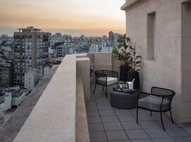 Um terraço no telhado com duas cadeiras de vime preto, uma mesa redonda pequena e um vaso de plantas.