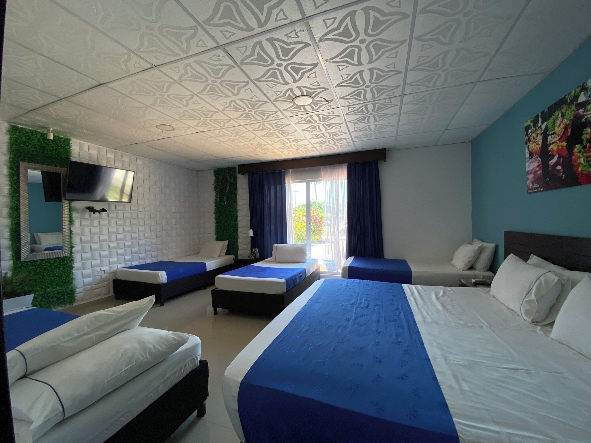 Quarto de hotel com uma cama grande, duas camas menores, uma televisão e teto com padrão geométrico.