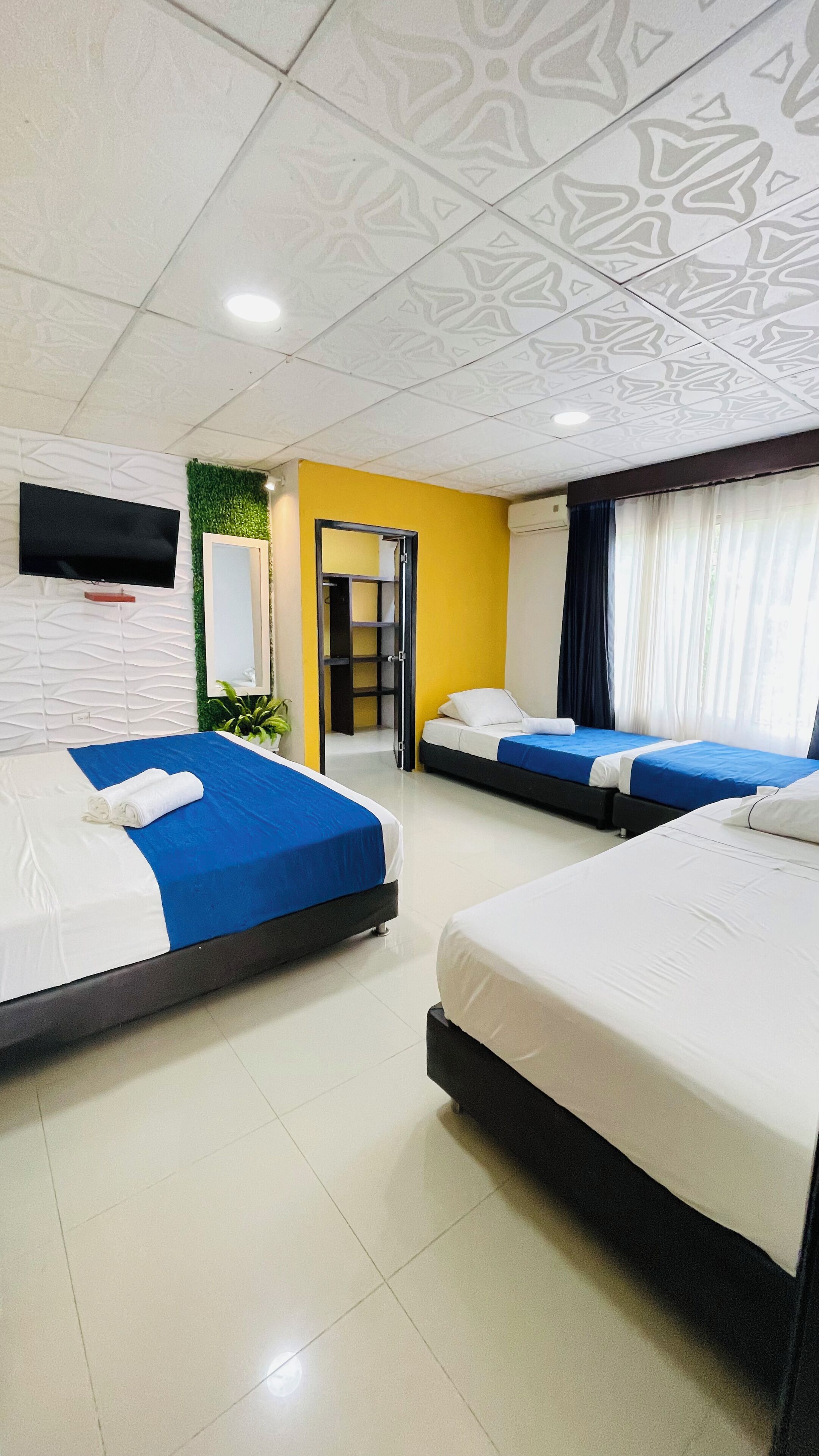 Quarto de hotel com três camas, televisão de tela plana fixada na parede, espelho e uma pequena planta.