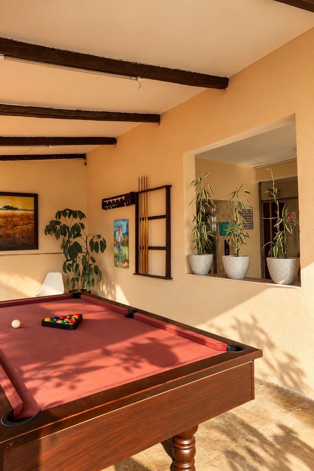 Mesa de bilhar com superfície de feltro vermelho, cercada por paredes com vigas de madeira, um quadro emoldurado e plantas em vasos.