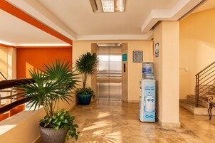 Corredor de hotel com bebedouro, plantas em vasos e elevador.