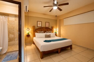 Quarto de hotel com cama, ventoinha no teto, área de chuveiro e ar condicionado fixado na parede.