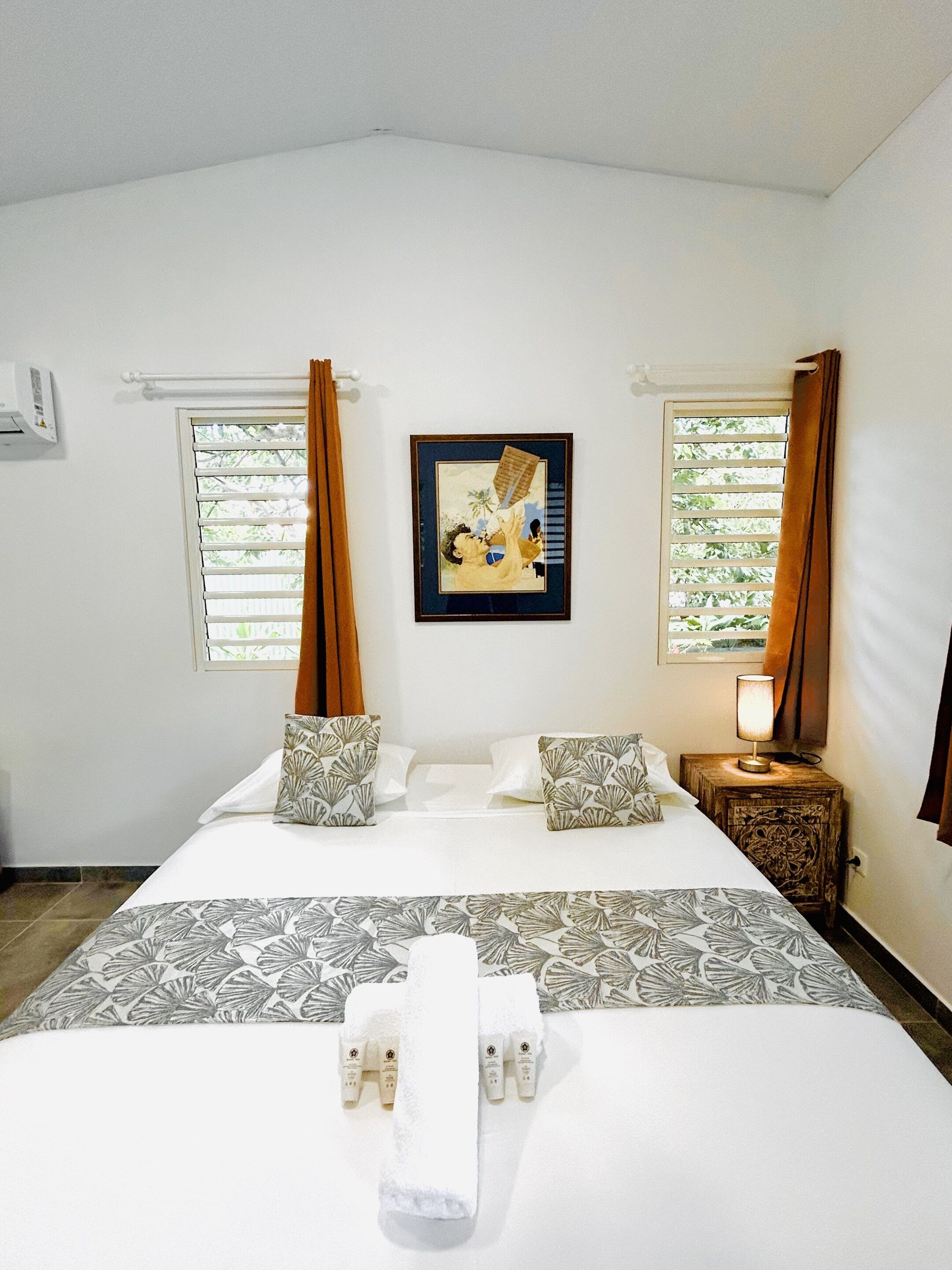 Quarto de hotel com cama, duas janelas com cortinas, um quadro na parede e uma mesa de cabeceira com abajur.