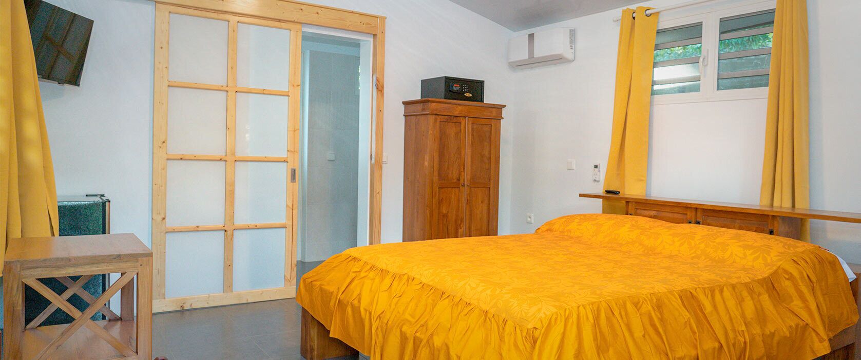 Quarto com cama, guarda-roupa de madeira, mesa pequena, televisão e janela com cortinas.