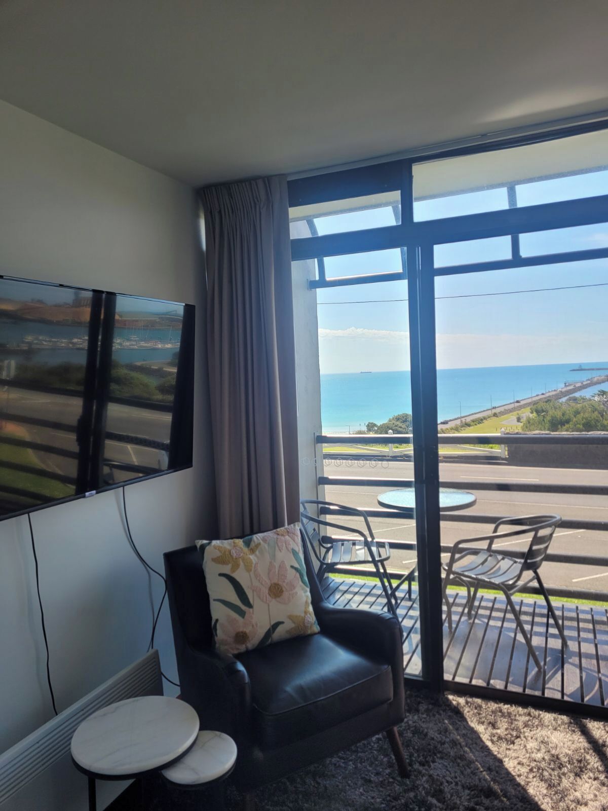 Quarto com uma grande janela que oferece vista para o mar e uma ponte ao longe. Há uma poltrona de couro preta com almofada floral, uma pequena mesa redonda e uma grande TV de tela plana presa à parede.