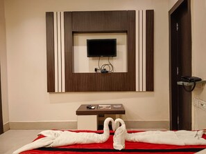 Quarto de hotel com televisão de tela plana na parede, uma mesinha com um controle remoto e uma revista, e um edredom de listras vermelhas e pretas.