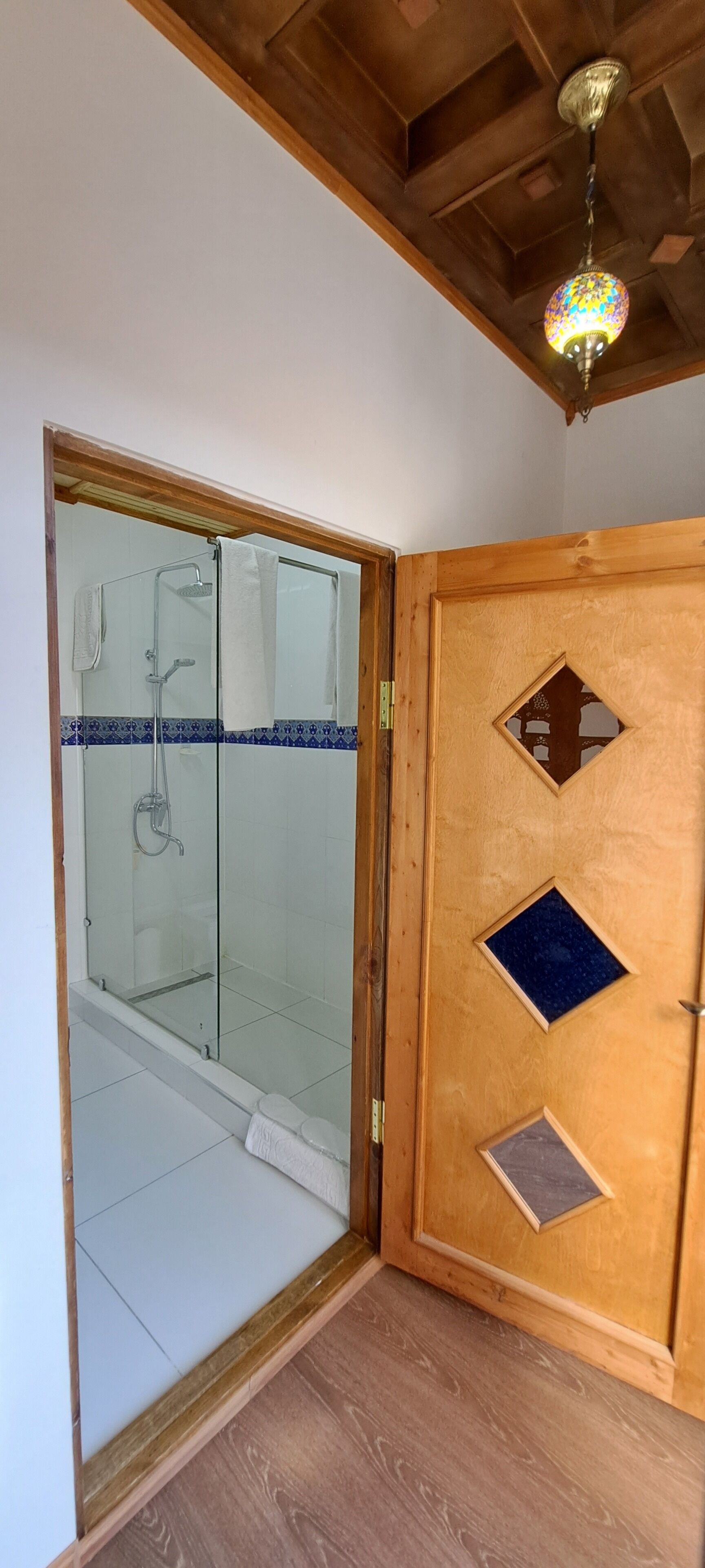 Quarto com chuveiro em cabine de vidro, porta de madeira com detalhes entalhados e luminária pendente.