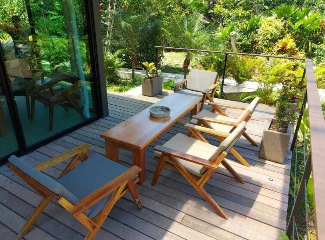 Um deck ao ar livre com mesa e cadeiras de madeira, cercado por vegetação exuberante.