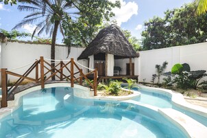 Uma piscina com uma ponte de madeira, uma estrutura com telhado de palha e plantas tropicais.