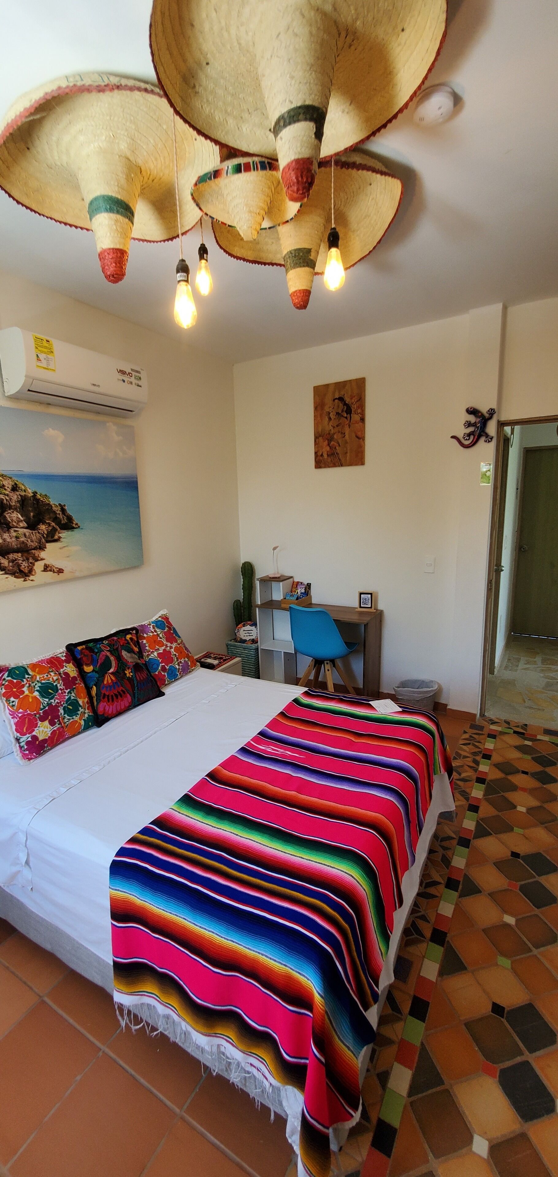 Quarto de hotel com cama, manta colorida, arte nas paredes e escrivaninha com cadeira.