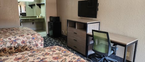 Quarto de hotel com cama, escrivaninha, cadeira, televisão e pequeno espaço de cozinha.