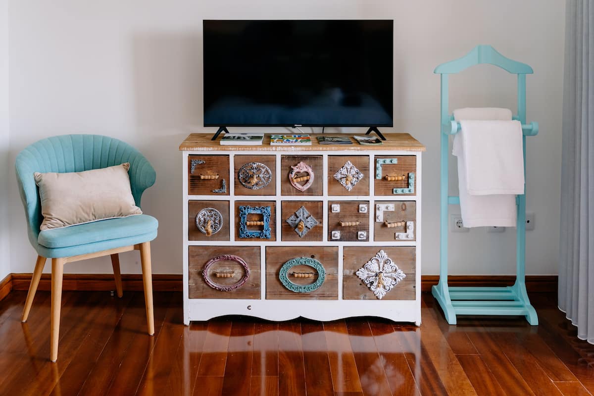 Quarto com um armário de madeira exibindo diversos objetos decorativos, uma poltrona verde-água, uma televisão e um porta-toalhas.