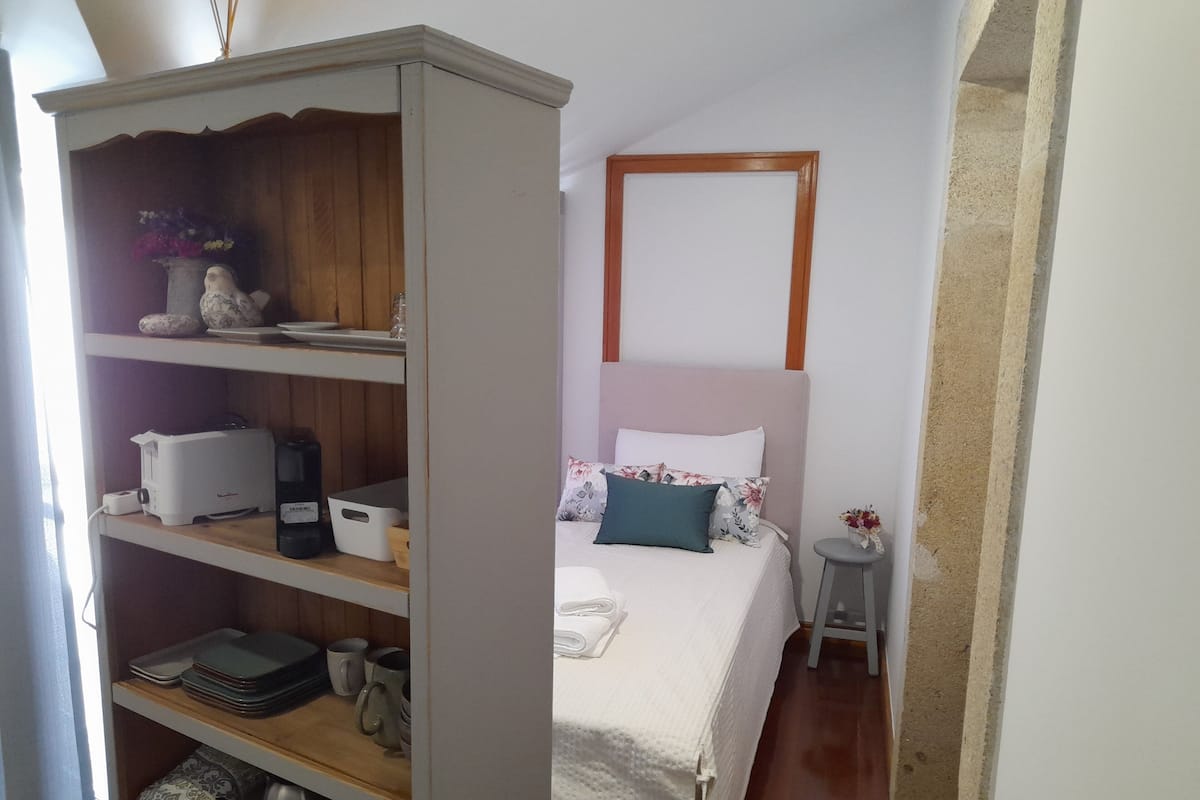 Quarto com cama, mesa de cabeceira, banquinho pequeno, guarda-roupa e prateleira com diversos objetos.