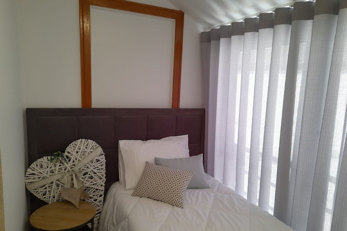 Quarto com cama grande, mesa de madeira redonda, cadeira branca decorativa e cortinas leves.