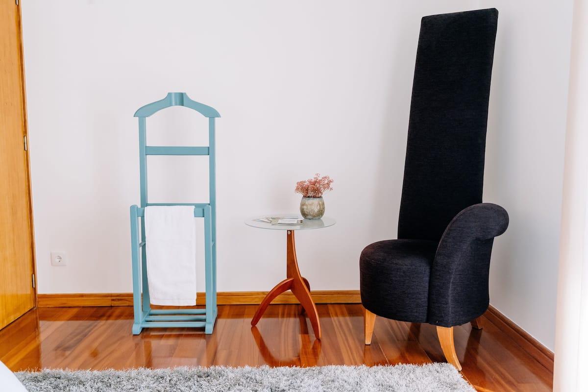 Quarto com piso de madeira, uma cadeira azul, um objeto preto alto, uma toalha branca em um suporte azul e uma pequena mesa com um vaso.