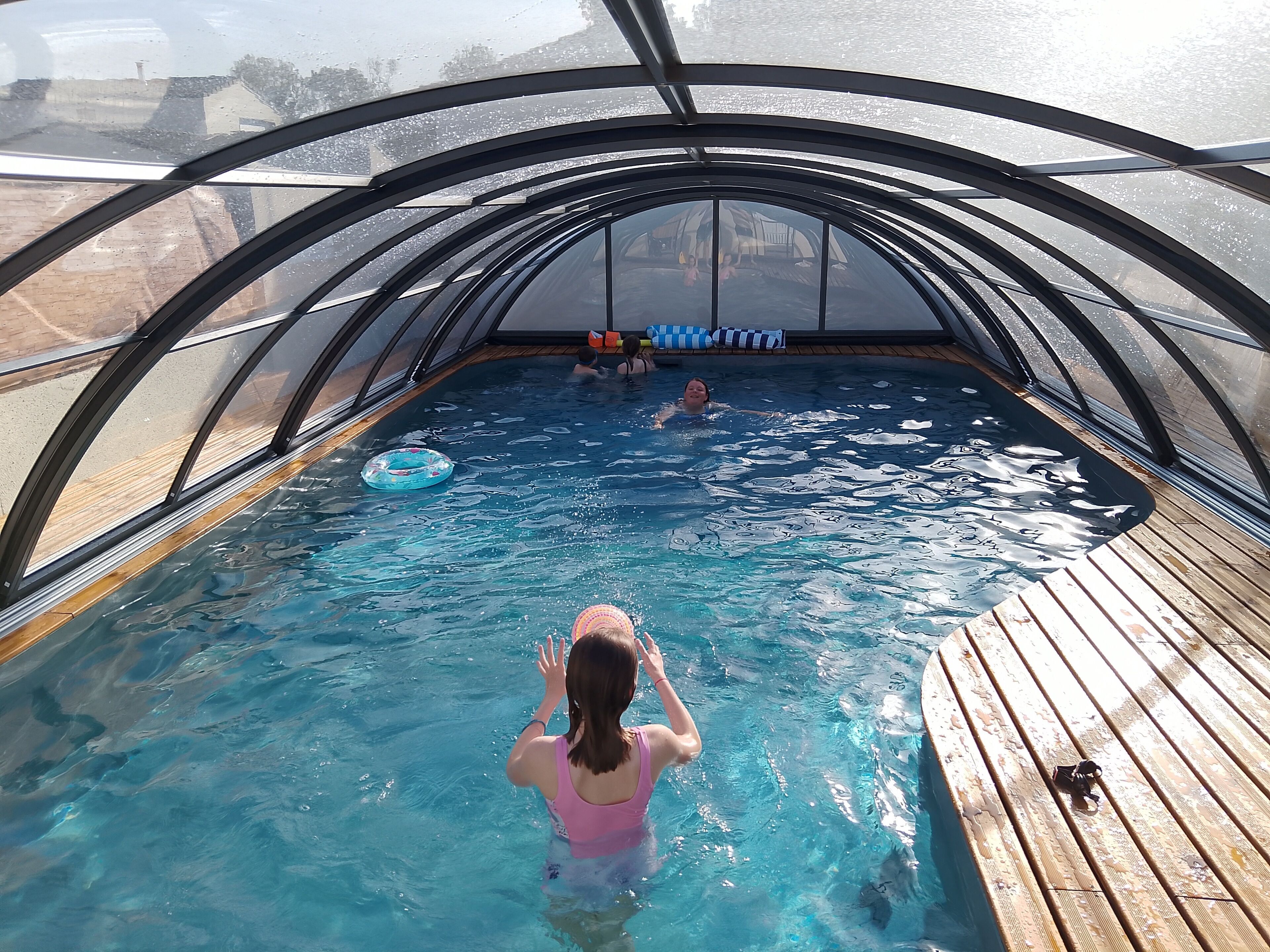 Uma piscina com cobertura transparente, deck de madeira e um flutuador.