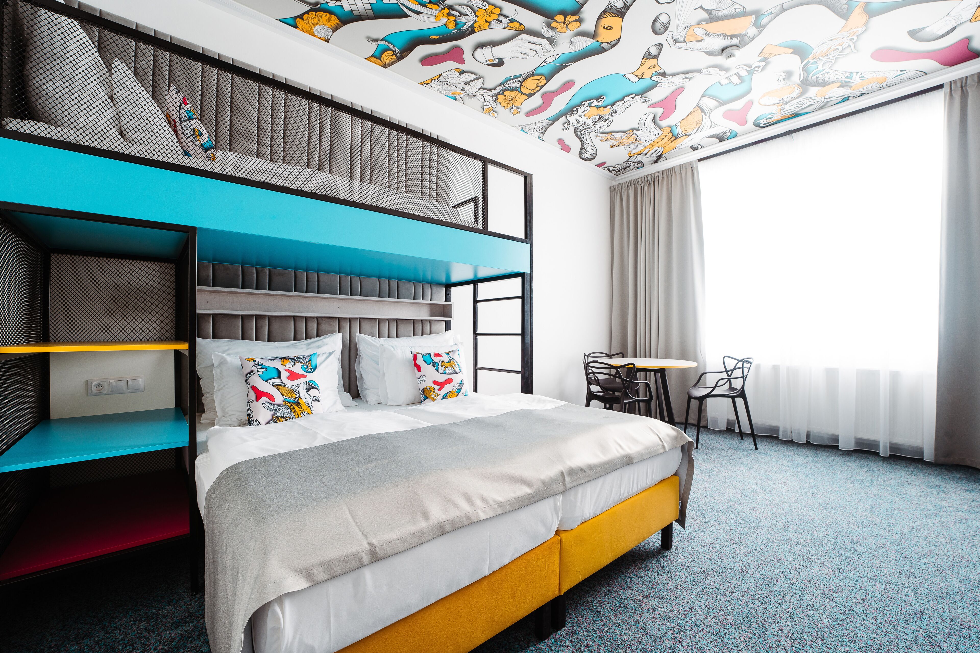 Quarto de hotel com beliche, escrivaninha, cadeira e uma janela grande com cortinas.
