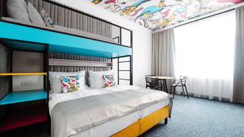 Quarto de hotel com beliche, escrivaninha, cadeira e uma janela grande com cortinas.