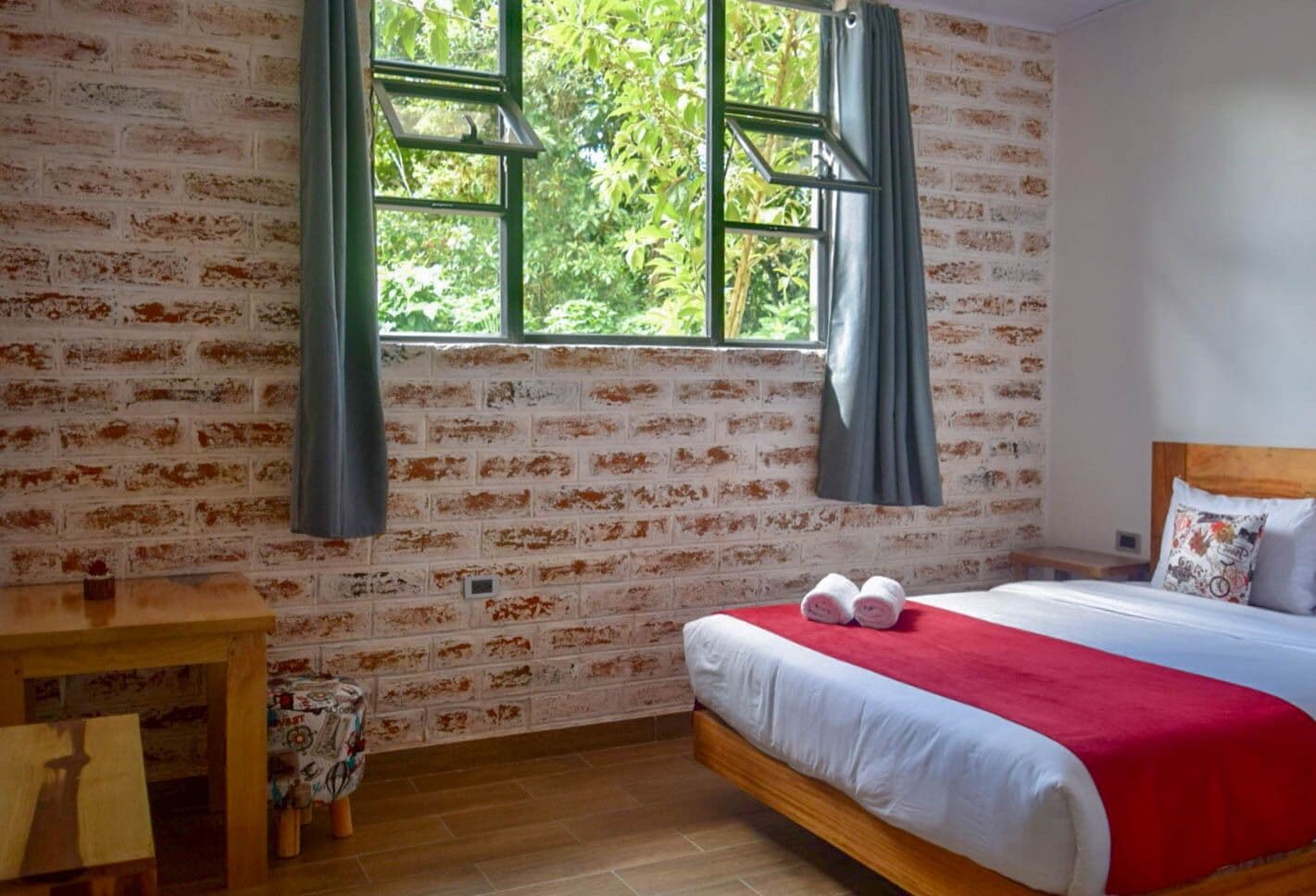 Quarto com parede de tijolos, cama de madeira com cobertor vermelho, mesa de cabeceira de madeira e janela com cortinas.