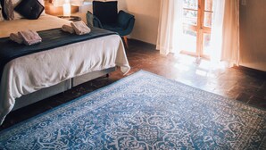 Quarto com uma cama, um tapete azul estampado, vigas de madeira no teto e uma janela com cortinas.