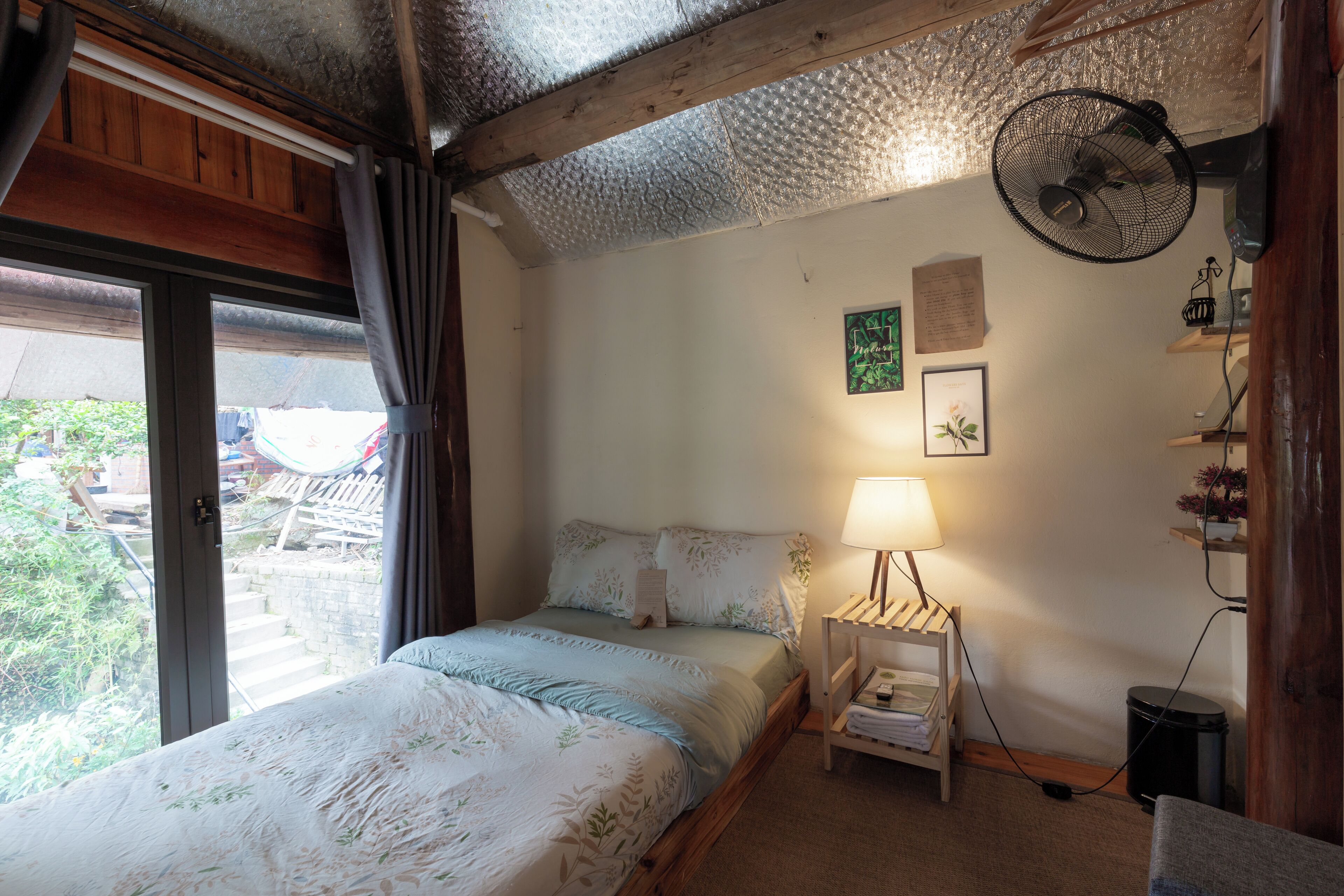 Quarto com cama, um ventilador, mesa de cabeceira com abajur e janela com cortinas.