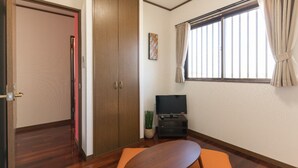 Quarto com chão de madeira, guarda-roupa de madeira, televisão sobre suporte, pequena mesa com almofadas laranja e janela com cortinas.