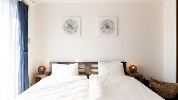 Uma cama de casal com lençóis brancos, duas luminárias fixadas na parede e duas obras de arte na parede.