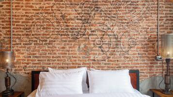 Quarto de hotel com parede de tijolo, uma cama com roupa de cama branca e duas luminárias em mesas de cabeceira de madeira.