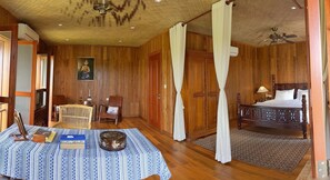 Um quarto com painéis de madeira, uma cama, uma mesa com toalha estampada, uma cadeira e um ventilador de teto.