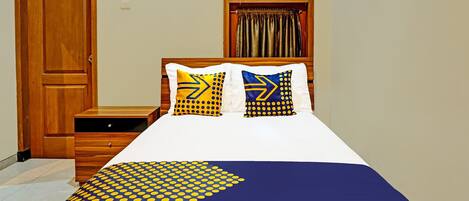 Uma cama bem arrumada com colcha azul e amarela, duas almofadas com padrão geométrico e uma mesinha de cabeceira de madeira.