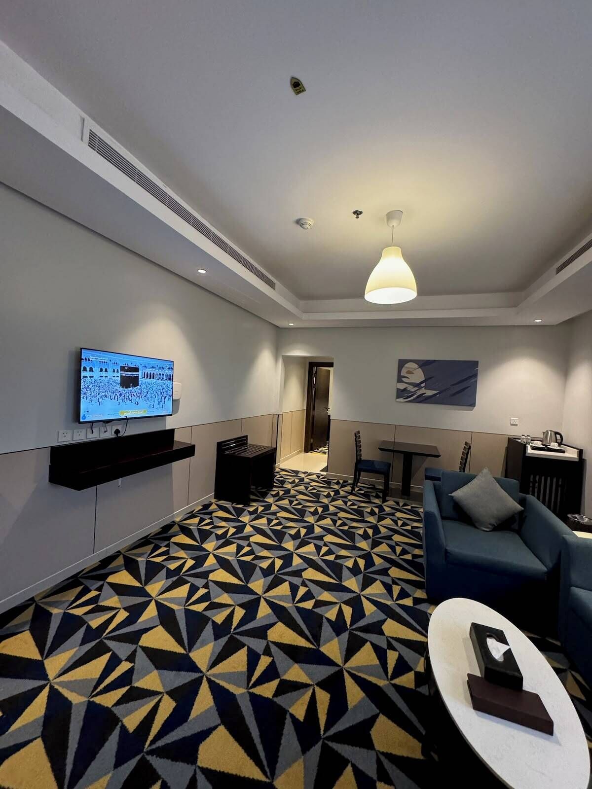 Quarto de hotel moderno com televisão de tela plana na parede, piso estampado, sofá azul, mesinha e luminária de teto.