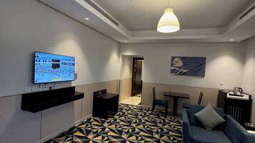 Quarto de hotel moderno com televisão de tela plana na parede, piso estampado, sofá azul, mesinha e luminária de teto.
