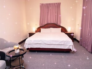 Quarto de hotel com uma cama, duas mesas de cabeceira com abajures, uma mesa redonda com um vaso de flores e um sofá cinzento.