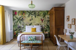 Quarto com cama, guarda-roupa de madeira, escrivaninha com cadeira, planta em vaso e parede com painel tropical.