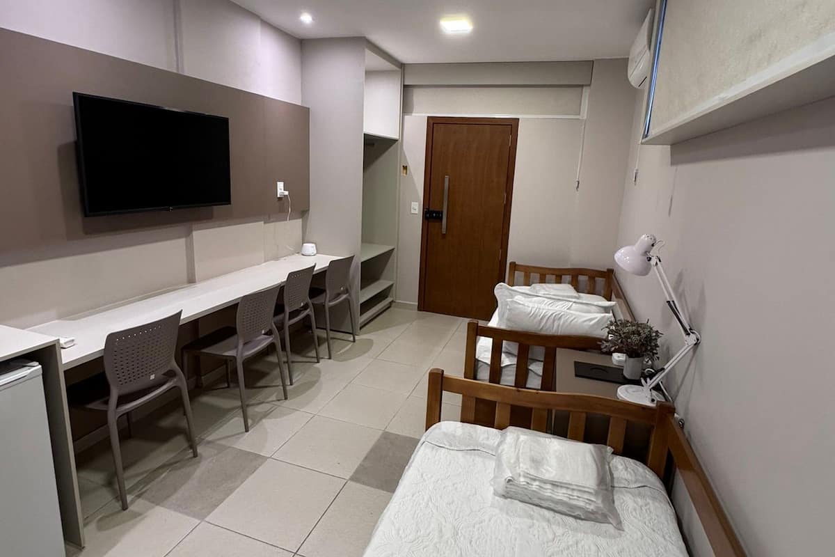 Quarto de hotel com cama, secretária com cadeiras, televisão e porta.