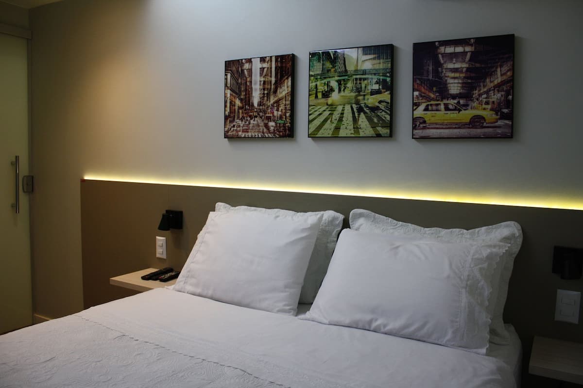 Quarto de hotel com uma cama, duas almofadas brancas, mesa de cabeceira com telefone e três quadros na parede.