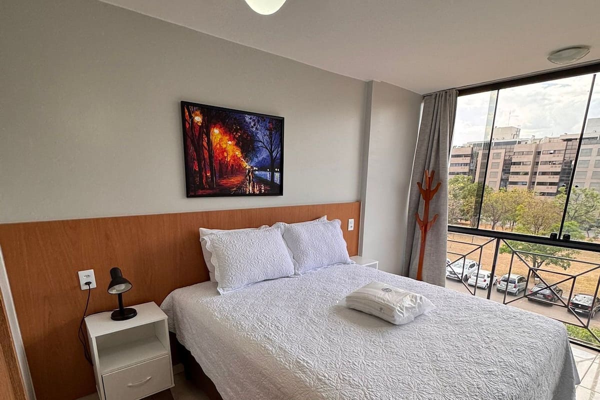 Quarto de hotel com uma cama, uma mesa de cabeceira, um abajur, um quadro na parede e vista da cidade por grandes janelas.