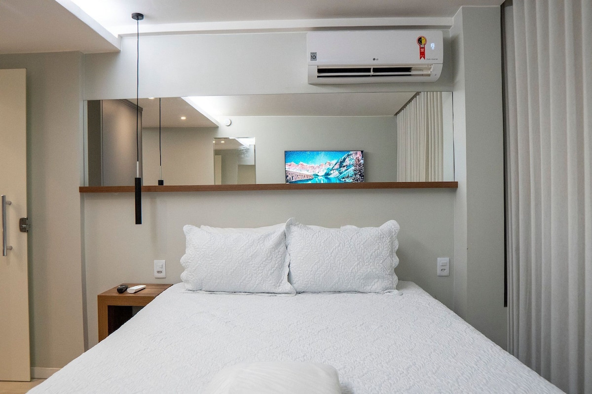 Quarto de hotel com cama, ar condicionado fixado na parede, um quadro e uma mesa de cabeceira com controle remoto.