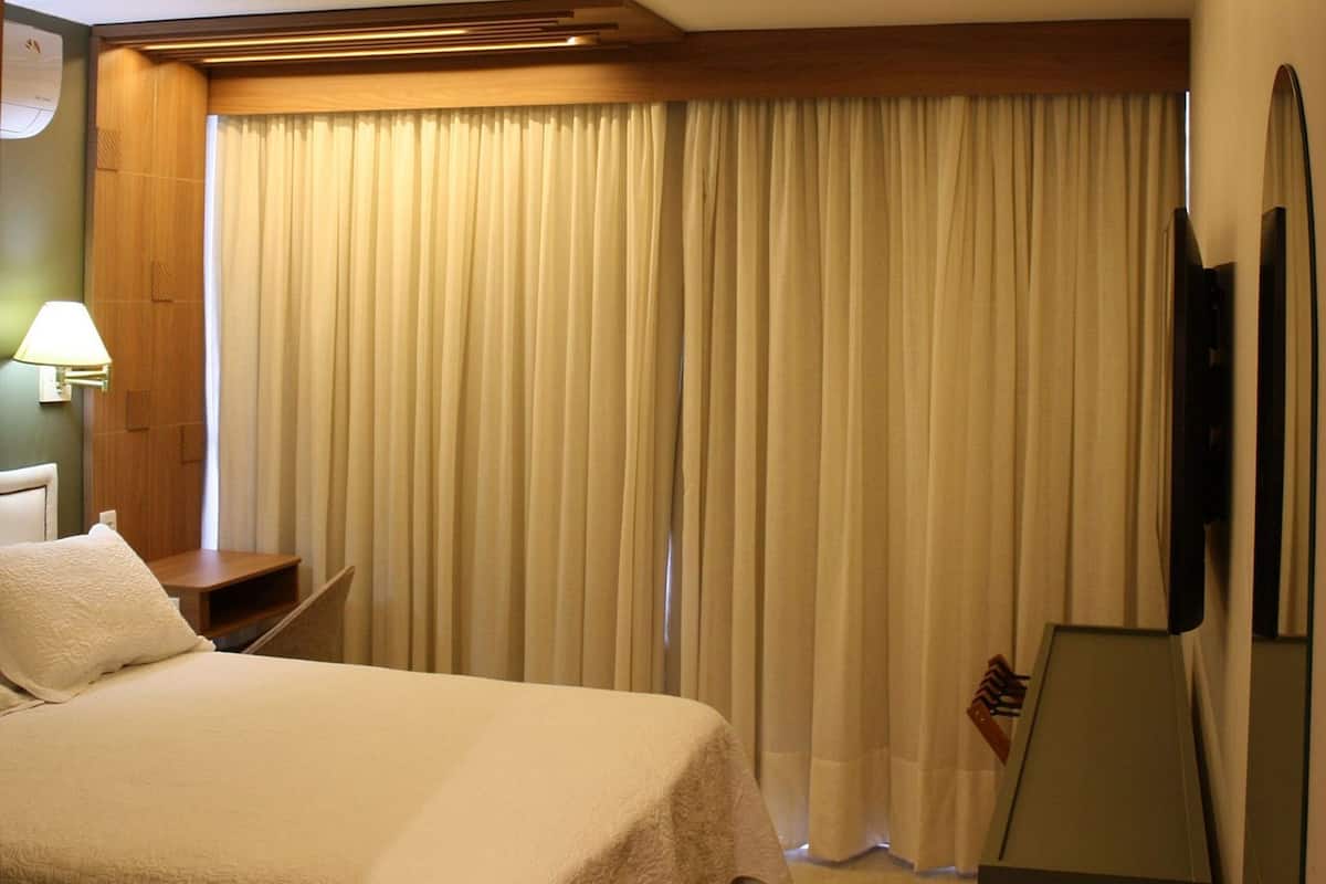 Quarto de hotel com cama, mesa de cabeceira, luminária, TV de tela plana e uma janela grande com cortinas.