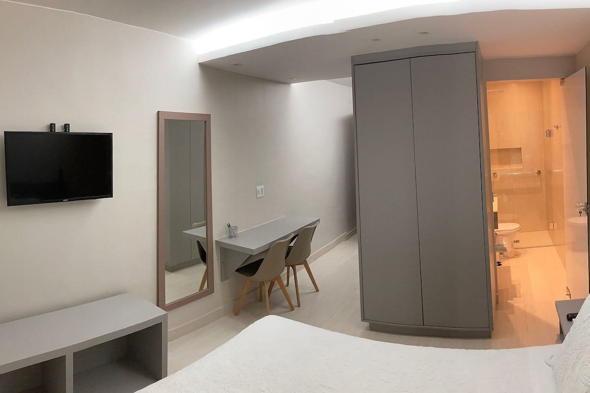 Quarto moderno com cama, escrivaninha, cadeira, guarda-roupa e televisão.