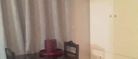 Um quarto pequeno com uma mesa de jantar e cadeiras, um guarda-roupa branco e uma cortina.