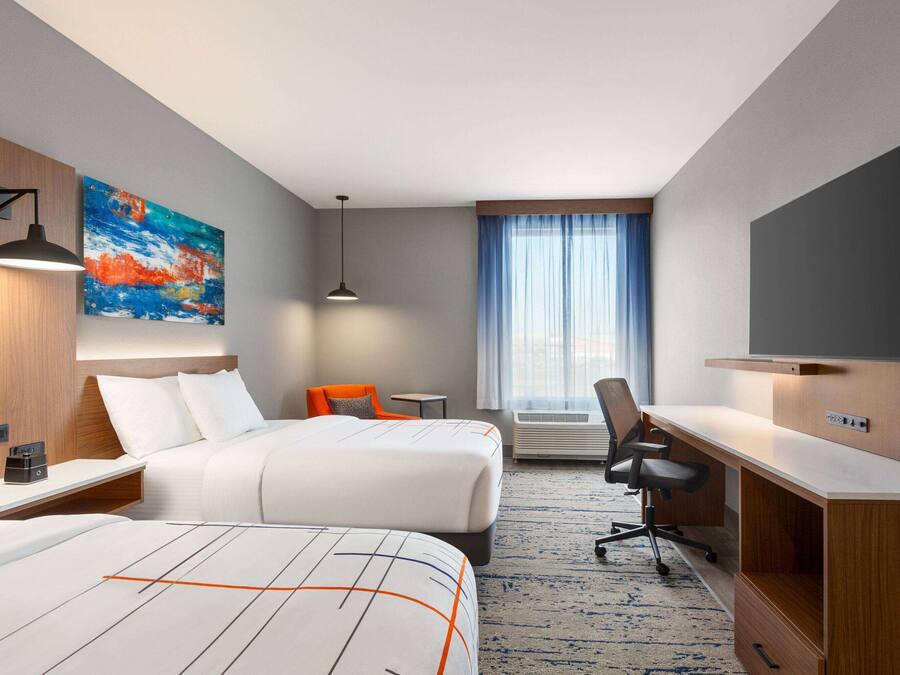Quarto de hotel com duas camas, uma escrivaninha, uma cadeira, uma televisão e um quadro na parede.