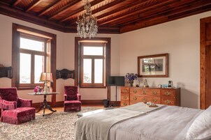 Quarto com uma cama grande, duas poltronas vermelhas, cômoda de madeira e um candelabro.