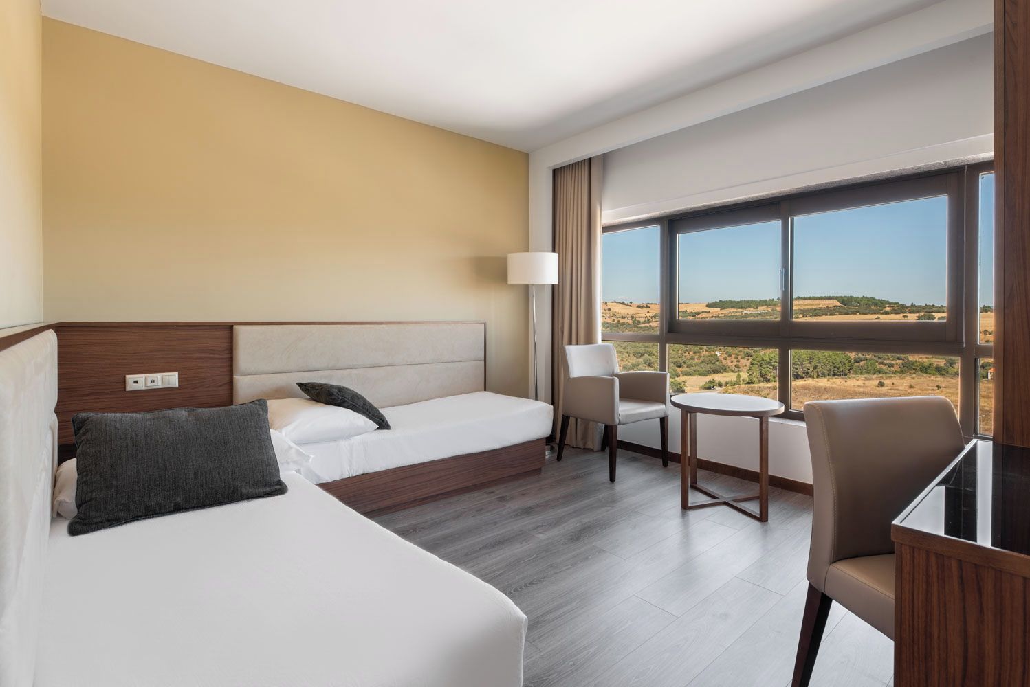 Quarto de hotel com duas camas, uma escrivaninha, uma cadeira e uma janela grande com vista para o campo.