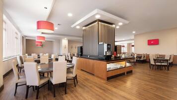 Um espaço de jantar moderno em hotel, com várias mesas e cadeiras, um ponto de café central e uma televisão de tela plana na parede.