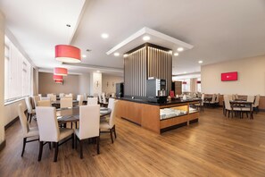 Um espaço de jantar moderno em hotel, com várias mesas e cadeiras, um ponto de café central e uma televisão de tela plana na parede.