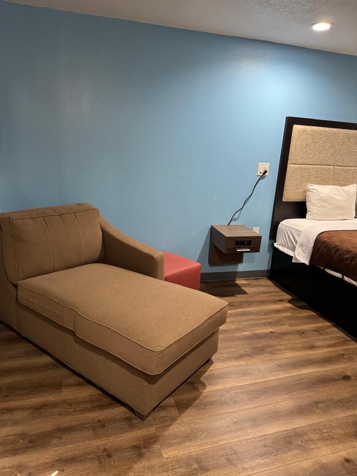 Um quarto com um sofá marrom, um banquinho vermelho pequeno, uma mesinha de cabeceira de madeira e uma cama com encosto.
