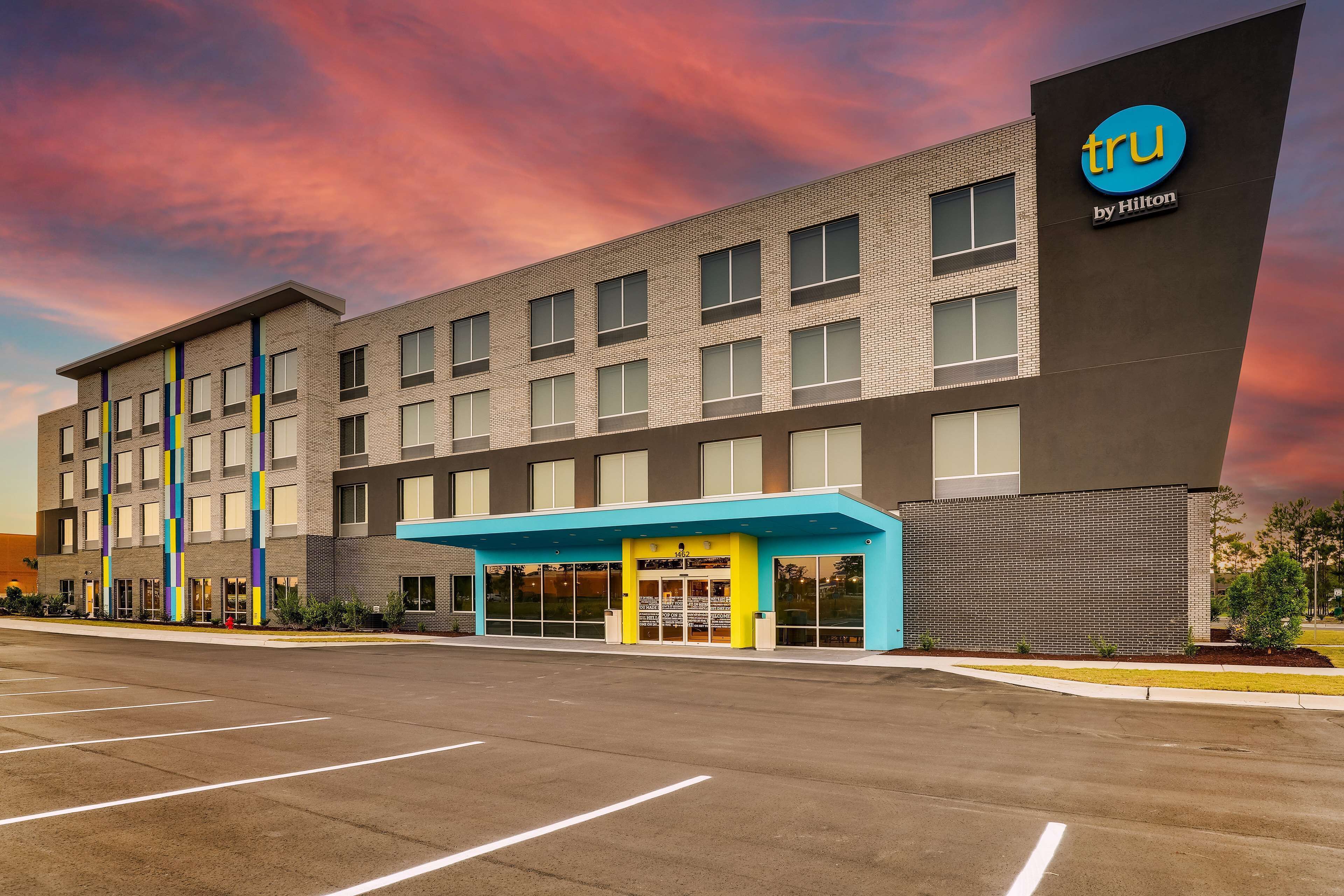 Um edifício de hotel moderno com um destaque para a placa 'tru by Hilton', uma cobertura de entrada azul e um estacionamento.