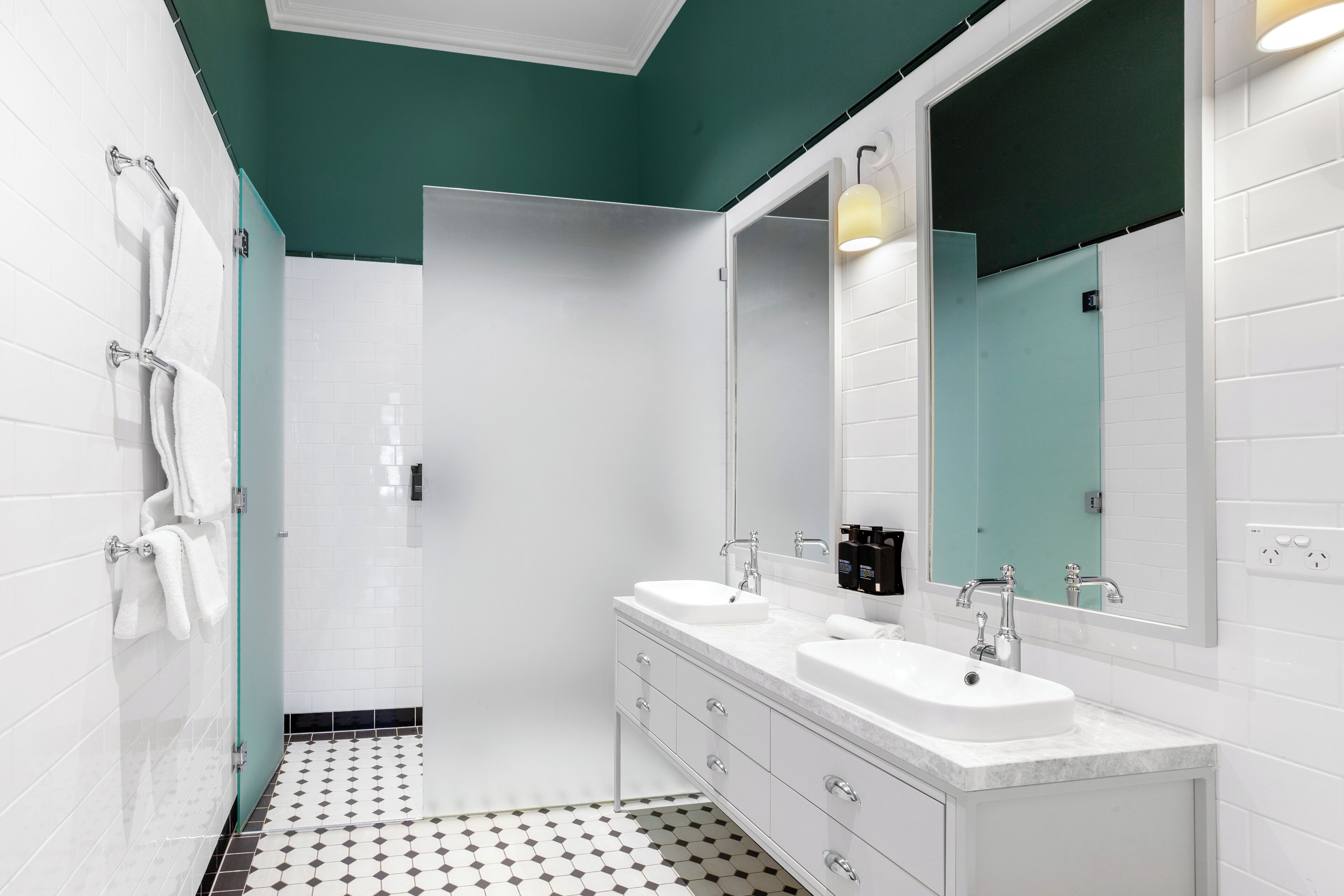 Um banheiro moderno com azulejos brancos, duas pias, um espelho grande e uma parede de destaque em tom verde-água.