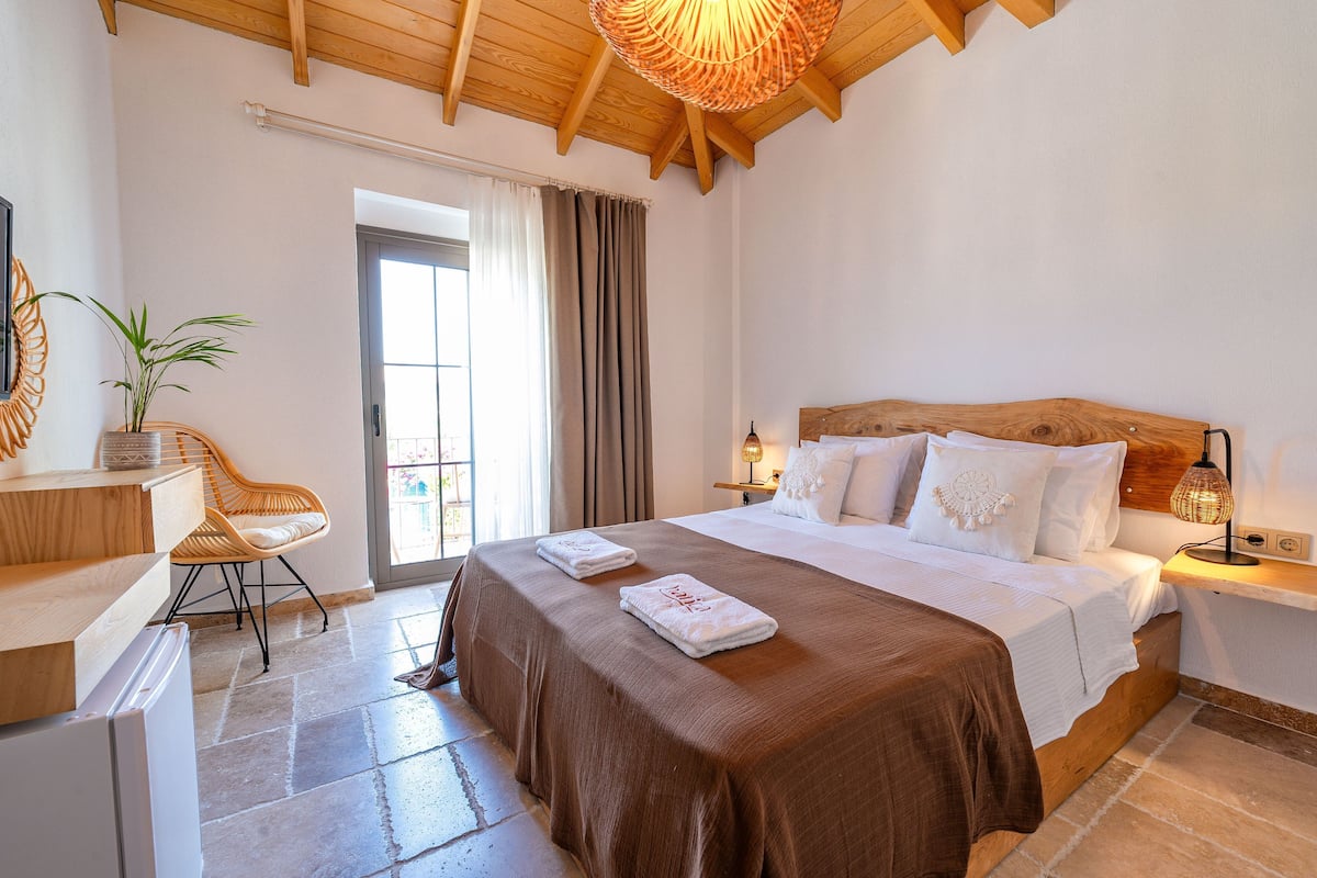 Quarto com cama grande, cabeceira de madeira, luminárias laterais, um vaso com planta e vista para o exterior pela janela.